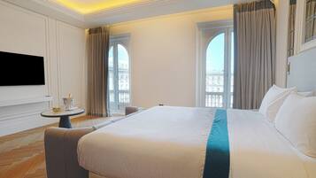 Quarto de hotel com uma cama grande, uma televisão, uma mesinha com uma bebida e vista para um prédio pela janela.