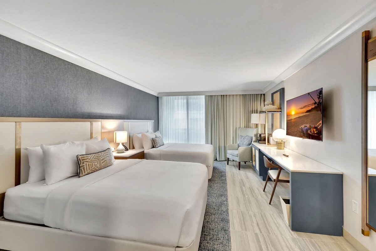 Quarto de hotel com duas camas, uma escrivaninha, uma cadeira, uma televisão e uma janela com cortinas.