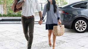 Um homem e uma mulher caminham de mãos dadas sobre um pavimento de azulejos, com um carro e palmeiras ao fundo.