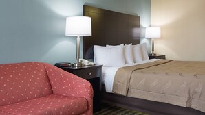 Quarto de hotel com cama, duas mesas de cabeceira com luminárias, um sofá vermelho estampado e cabeceira.