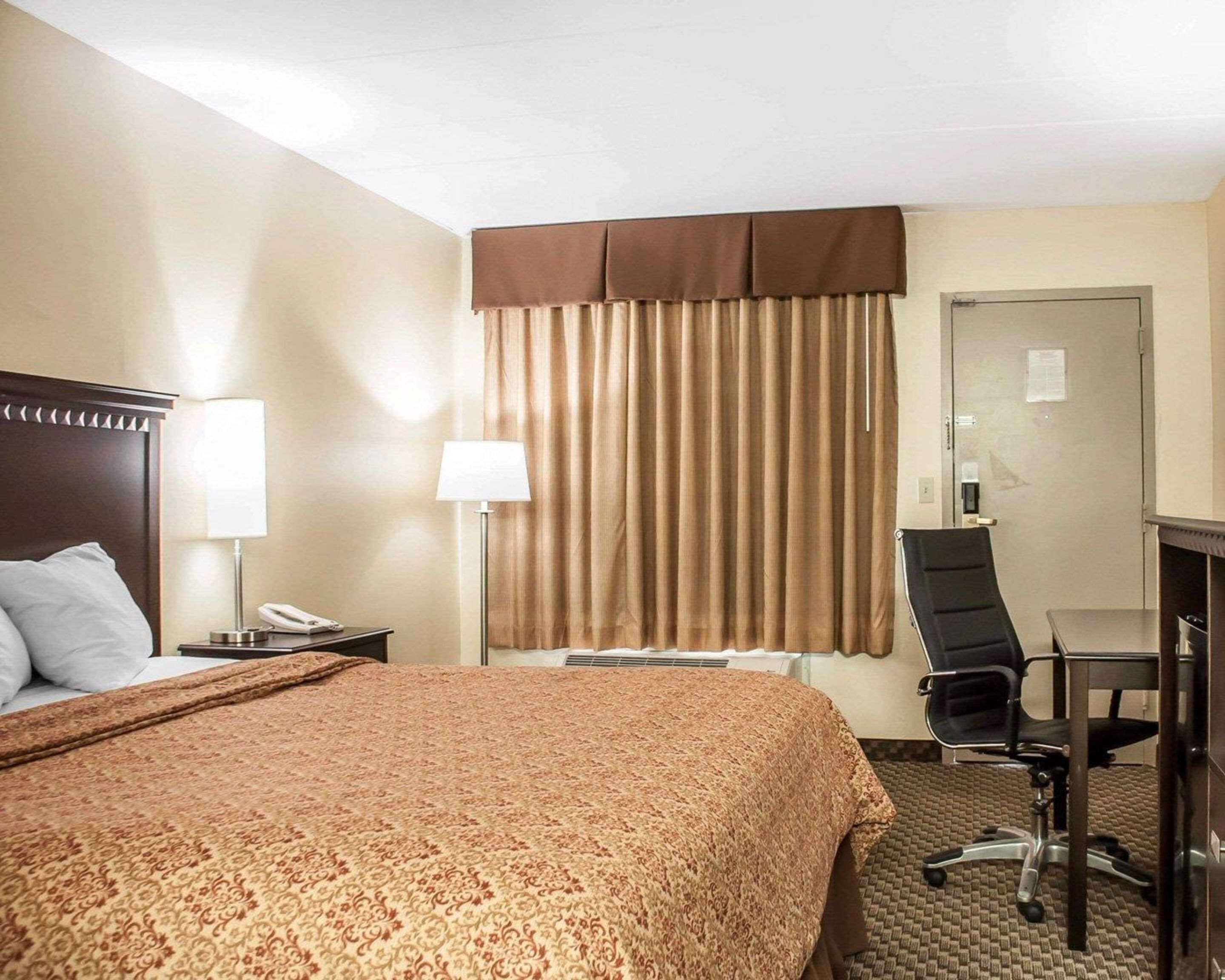 Quarto de hotel com uma cama grande, uma mesa com cadeira, uma luminária e uma janela com cortinas.