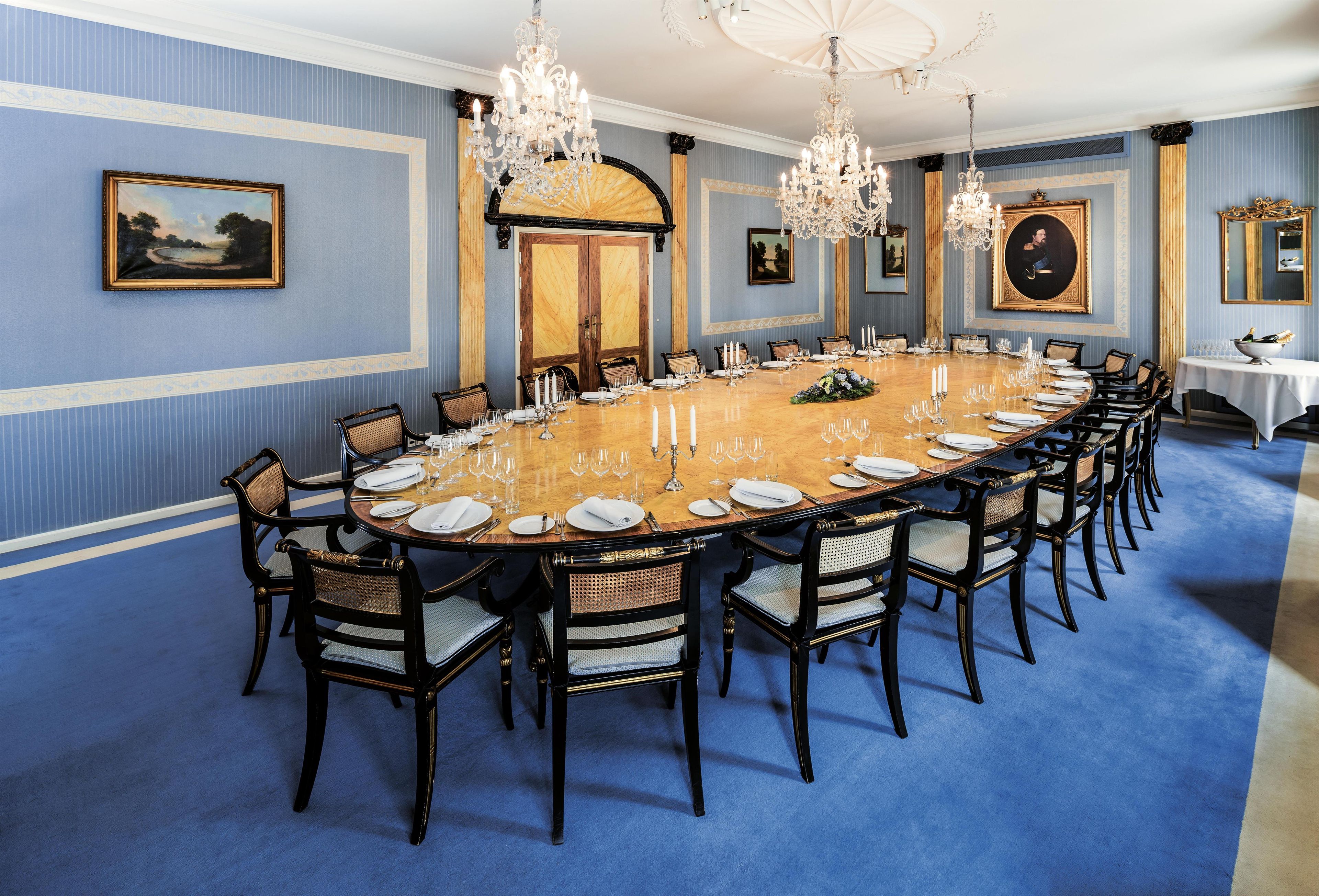 Uma sala de jantar formal com uma grande mesa oval, cadeiras ricamente decoradas e lustres.