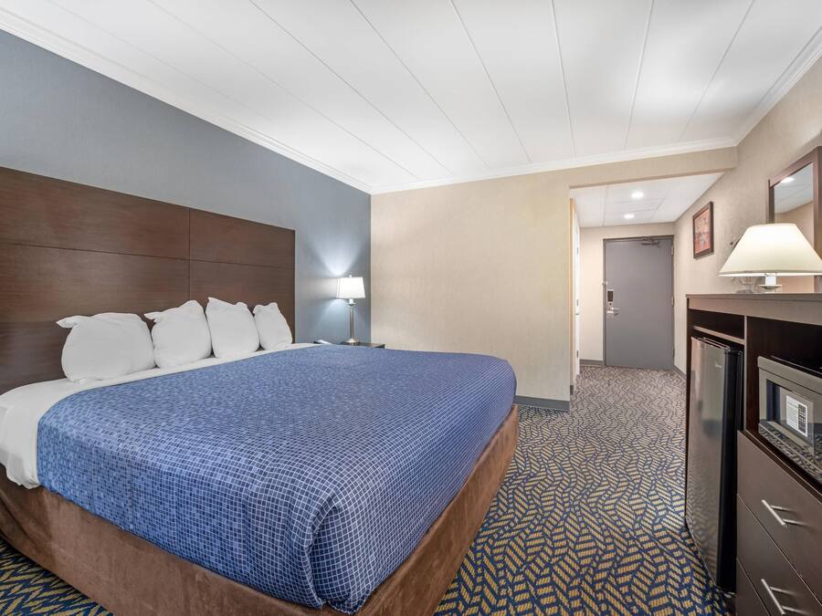 Quarto de hotel com uma cama grande, uma mesa com luminária, um minibar e uma porta que dá para outro cômodo.