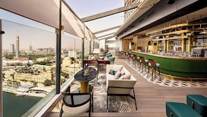 Um bar no terraço com vista para a cidade, área de estar com cadeiras e um sofá, e balcão de bar com garrafas.