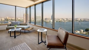 Um quarto amplo com uma grande janela que oferece vista para o horizonte urbano e um corpo d'água. Há um sofá, uma poltrona e uma pequena mesa com uma planta.