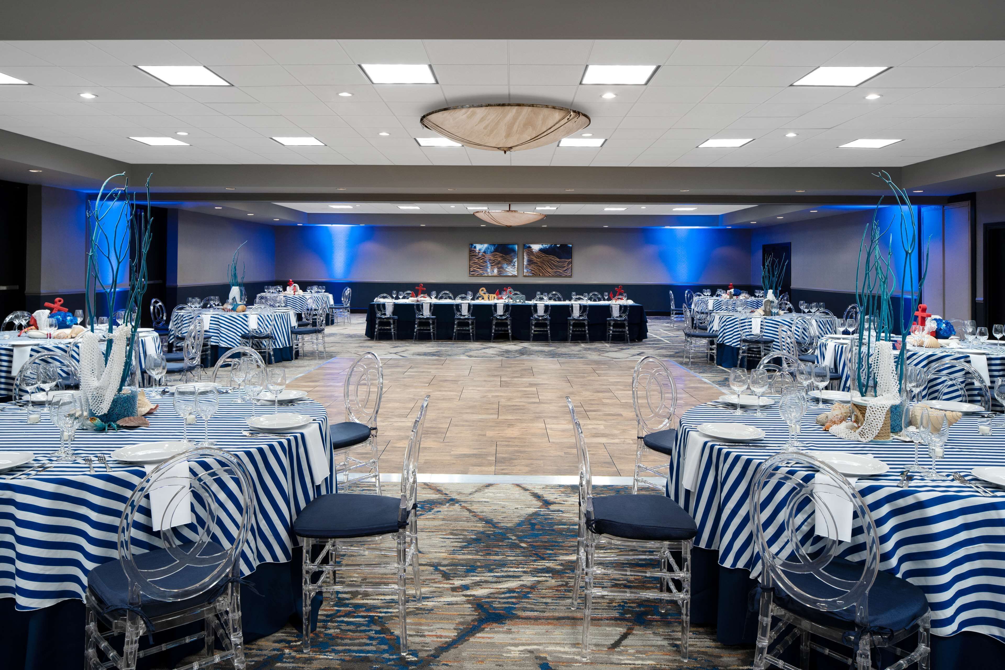 Uma sala de banquetes com mesas montadas para um evento formal, com cadeiras transparentes e toalhas de mesa listradas em azul.