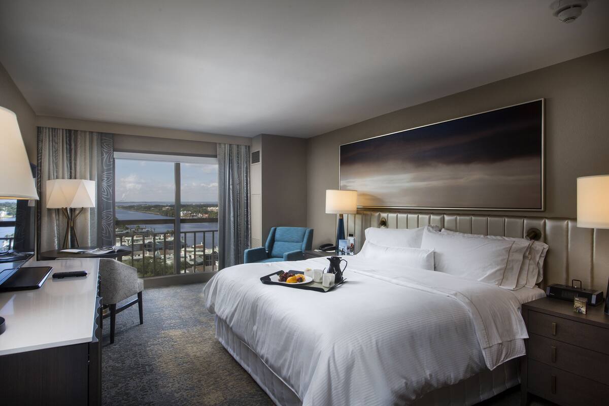 Quarto de hotel com uma cama grande, uma escrivaninha com um laptop, uma cadeira e uma bandeja com frutas e uma xícara.