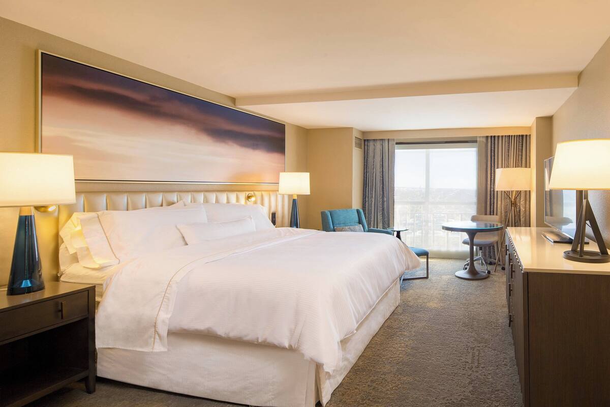 Quarto de hotel com uma cama grande, duas mesas de cabeceira com abajures, uma cadeira, uma escrivaninha com luminária e uma janela grande com cortinas.