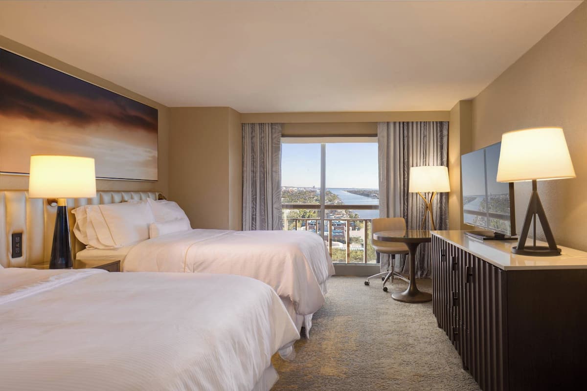 Quarto de hotel com duas camas, uma escrivaninha, uma cadeira, uma televisão e uma janela grande com vista para a paisagem urbana.