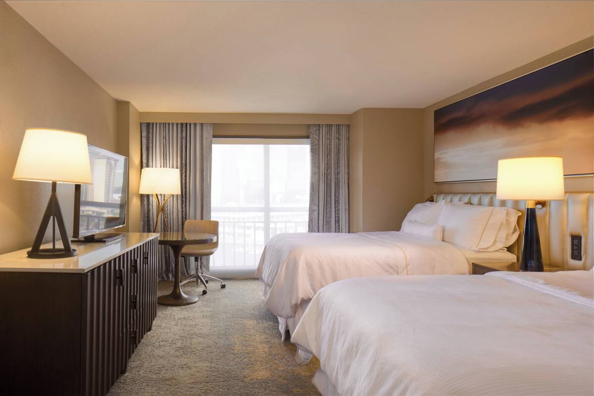 Quarto de hotel com duas camas, uma mesa de trabalho com televisão e uma janela grande com cortinas.