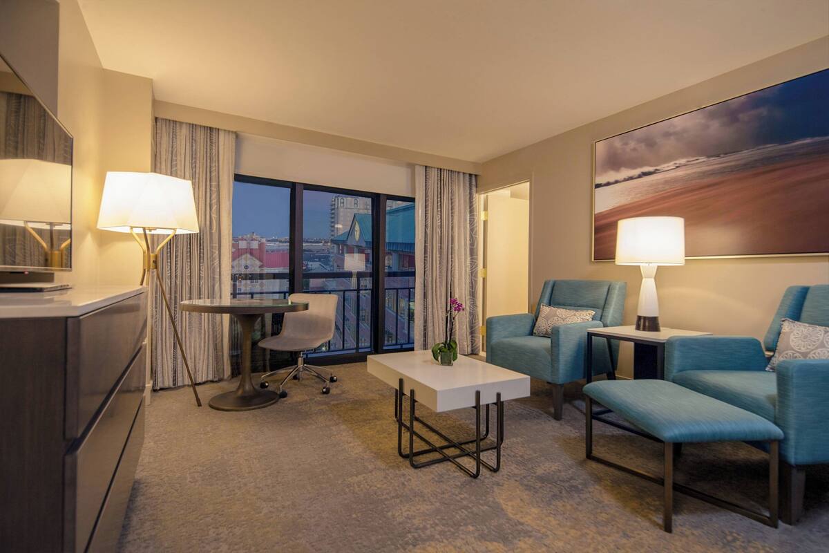 Quarto de hotel com uma janela grande, uma escrivaninha, duas poltronas, uma mesinha e um quadro na parede.
