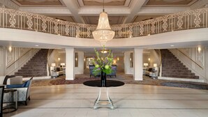 Um amplo hall de entrada com um candelabro, uma escadaria e uma mesa central com arranjo de flores.