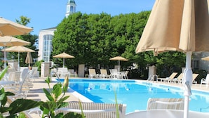 Uma área de piscina com espreguiçadeiras e guarda-sóis, uma piscina azul cristalina e um edifício com cúpula ao fundo.