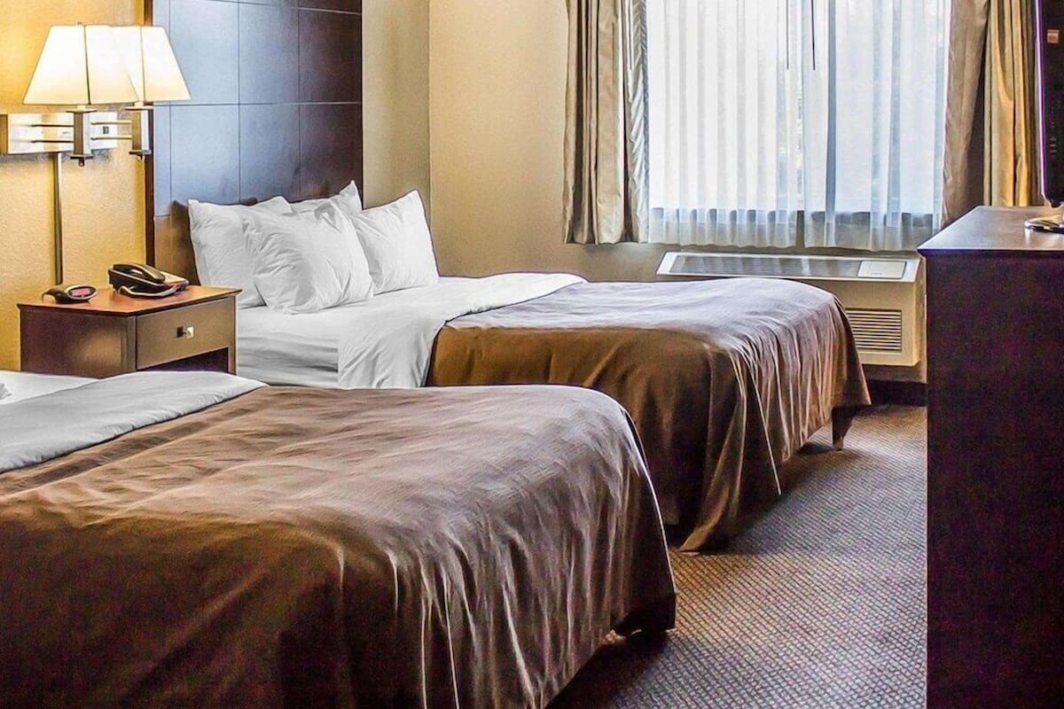 Quarto de hotel com duas camas, mesa de cabeceira com abajur, escrivaninha com computador e janela com cortinas.