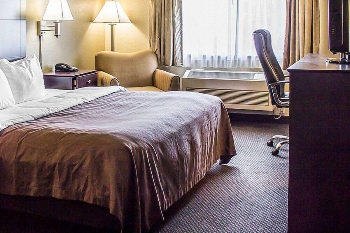 Quarto de hotel com uma cama grande, duas cadeiras, uma escrivaninha, um abajur, um telefone e uma televisão.