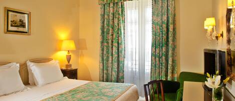 Quarto de hotel com cama, escrivaninha, cadeira, abajur, um quadro na parede e janela com cortinas.