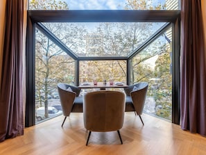Uma área de refeições com mesa e cadeiras, grandes janelas e vista para árvores e edifícios.