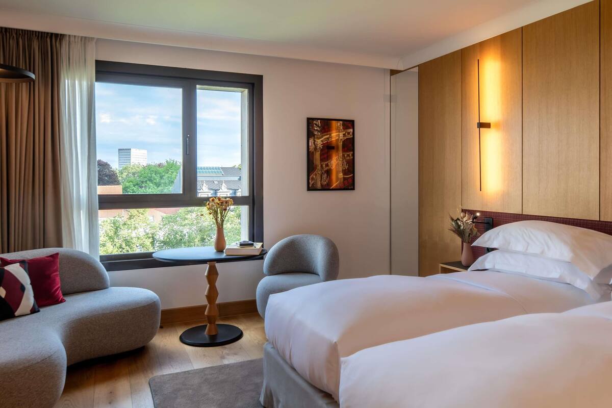 Quarto de hotel com cama, sofá, cadeira, pequena mesa com um vaso e um quadro na parede.