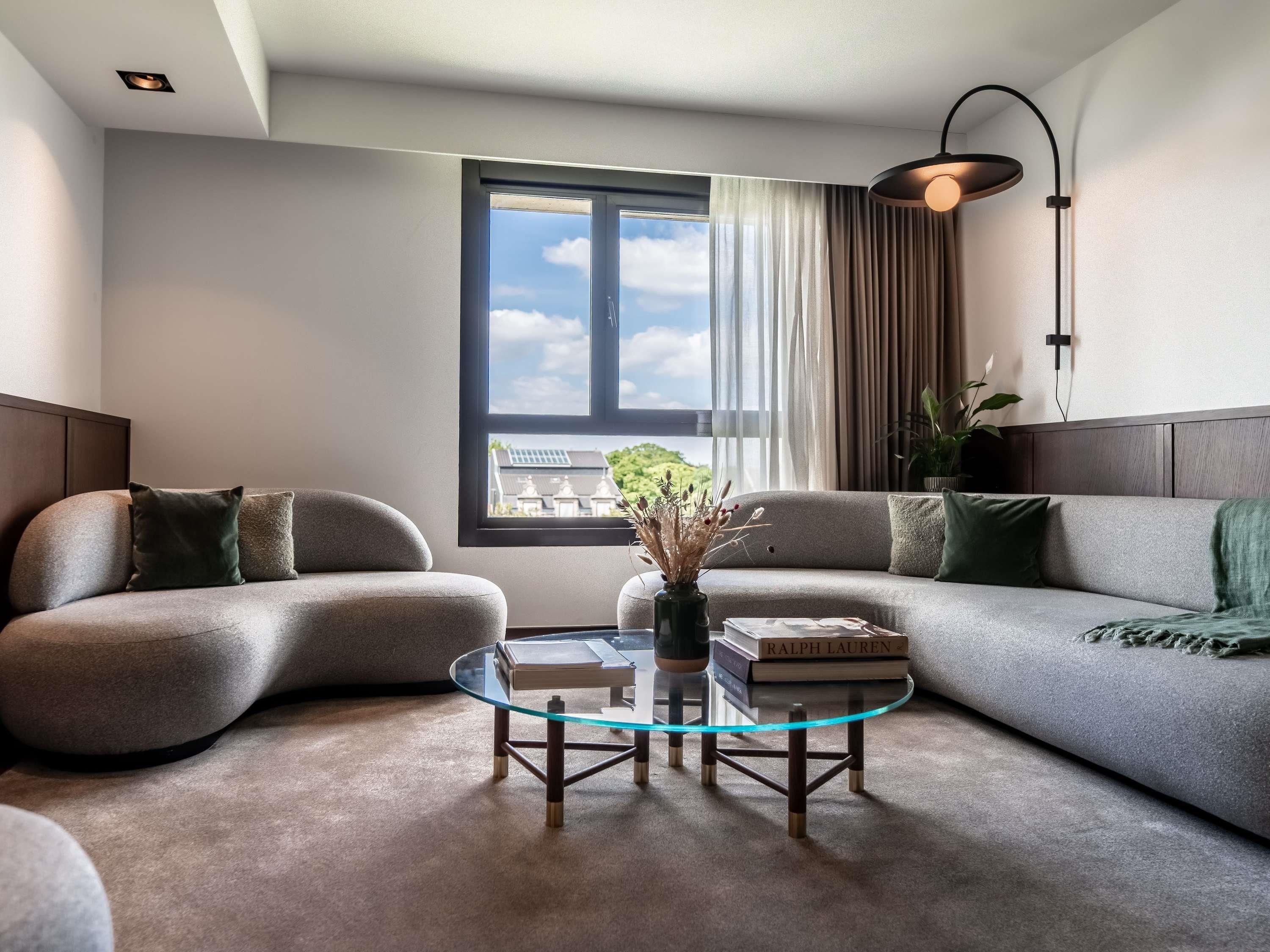 Uma sala de estar moderna com uma mesa de centro de vidro, dois sofás, uma janela grande com cortinas e um abajur.