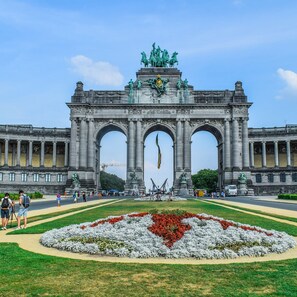Um grande arco histórico com estátuas e um emblema central, rodeado por um canteiro circular de flores.