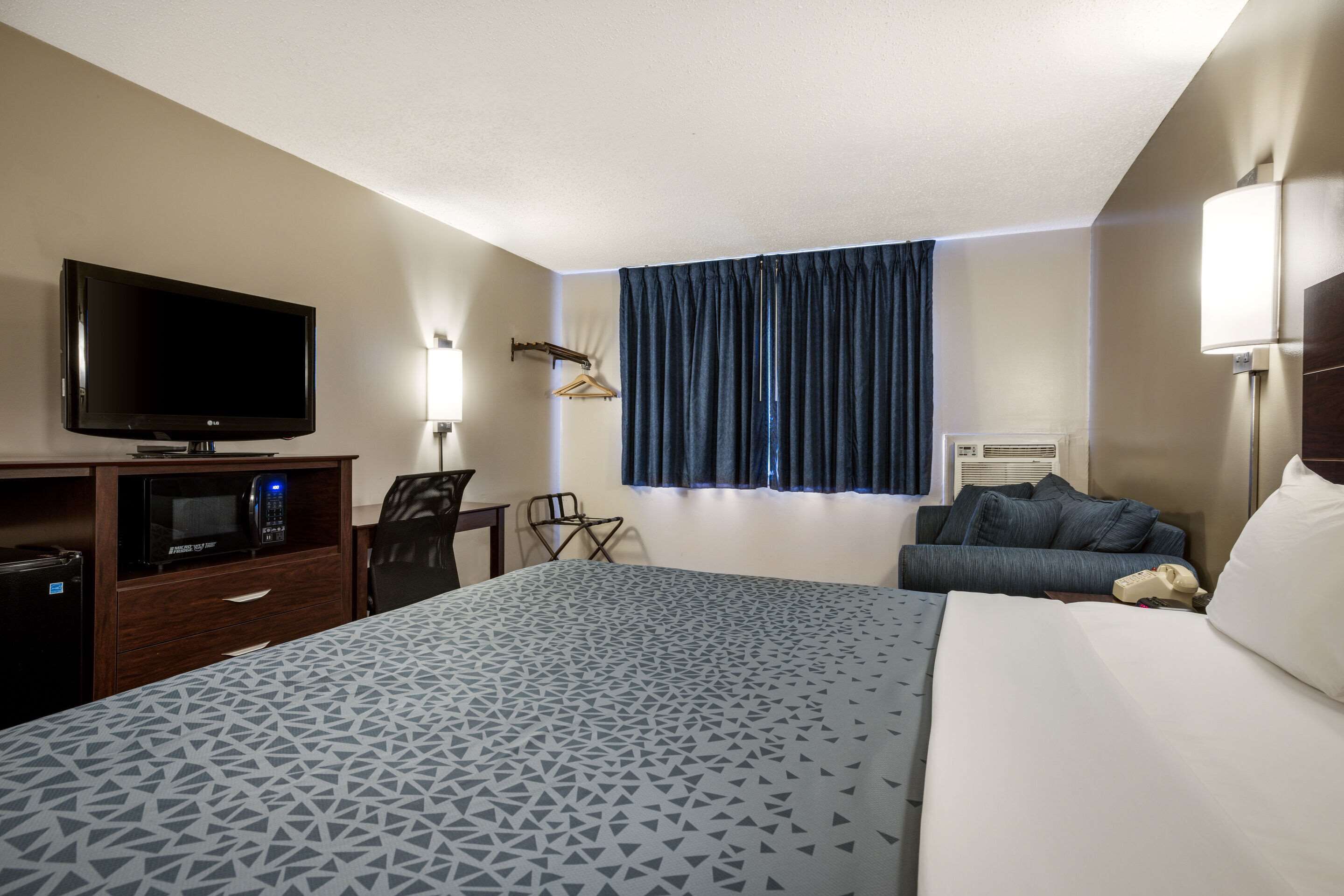 Quarto de hotel com cama, televisão, secretária com computador, cadeira e janela com cortinas.