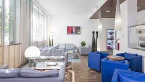 Uma sala de estar moderna com um sofá cinzento, poltronas azuis e uma televisão fixada na parede.
