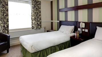Quarto de hotel com duas camas de solteiro, cabeceira de madeira, mesa de cabeceira com abajur e janela com cortinas.