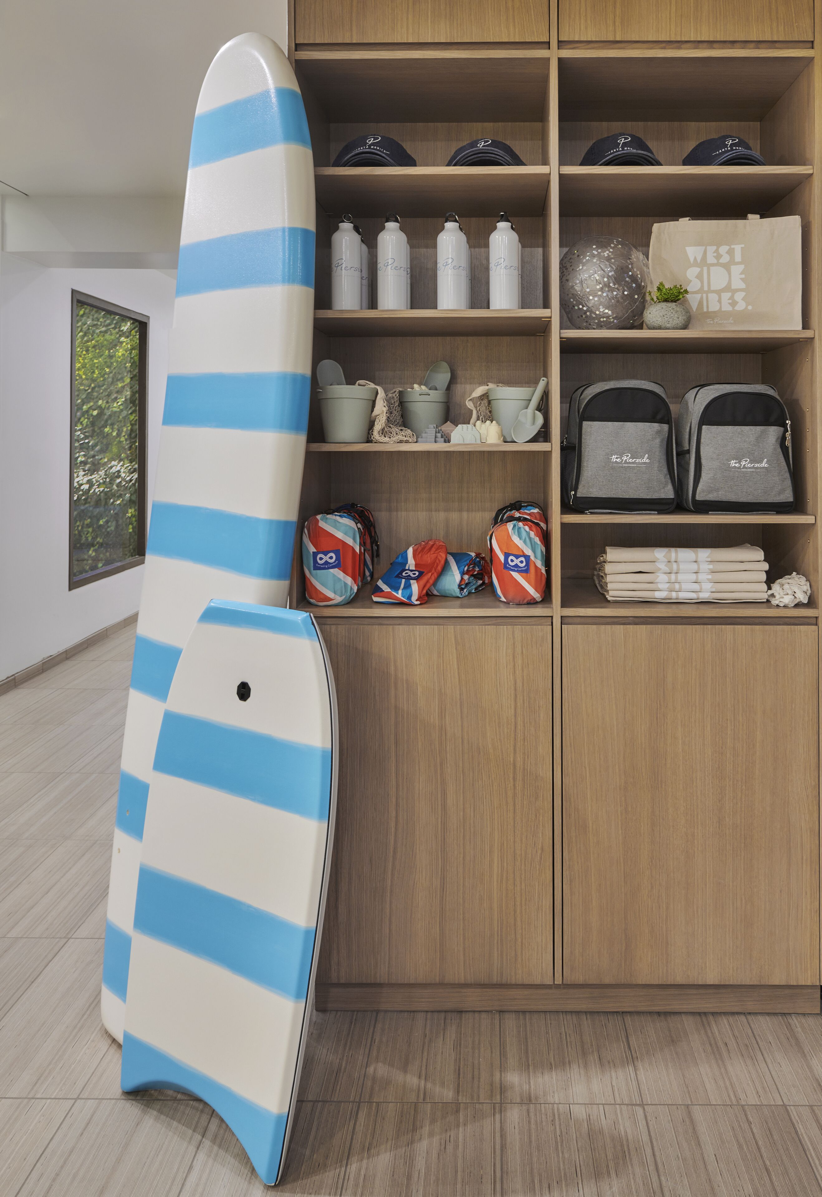 Quarto com uma prancha de surf apoiada em um armário de madeira, janela com vista para árvores e prateleiras com diversos objetos, incluindo bolsas e garrafas.