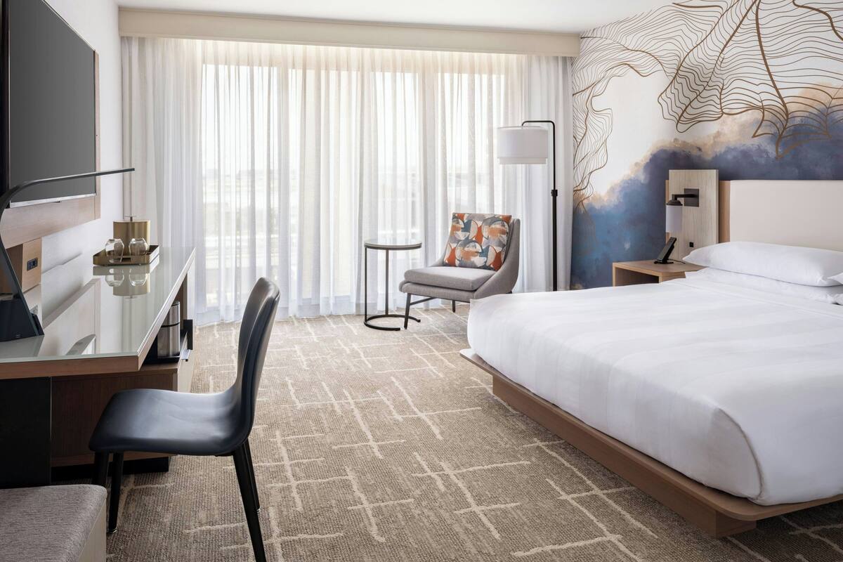 Quarto de hotel com uma cama grande, uma escrivaninha com cadeira, uma mesinha e um abajur.