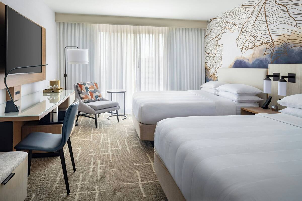 Quarto de hotel com duas camas, uma escrivaninha com cadeira, televisão e uma janela grande com cortinas.