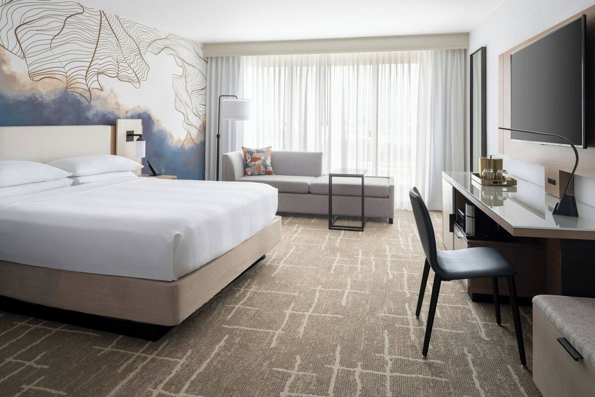 Quarto de hotel com uma cama grande, um sofá, uma escrivaninha com cadeira e uma televisão.