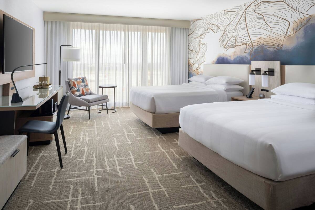 Quarto de hotel com duas camas, uma escrivaninha com cadeira, televisão e uma janela grande com cortinas.