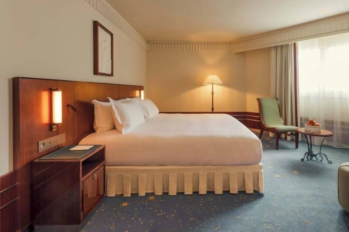 Quarto de hotel com cama grande, mesa de cabeceira, abajur, cadeira e uma pequena mesa com um bolo.