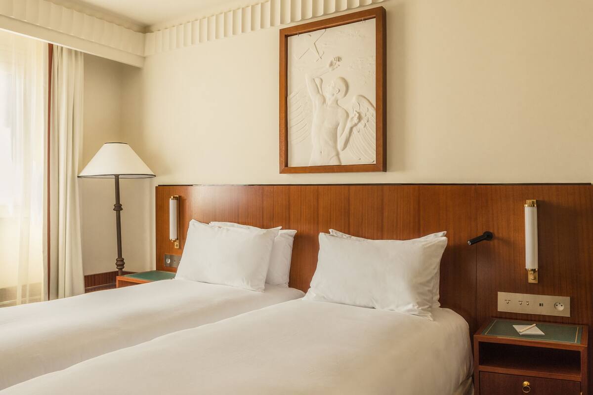 Quarto de hotel com duas camas, uma mesa de cabeceira com abajur, uma obra de arte na parede e cabeceira de madeira.