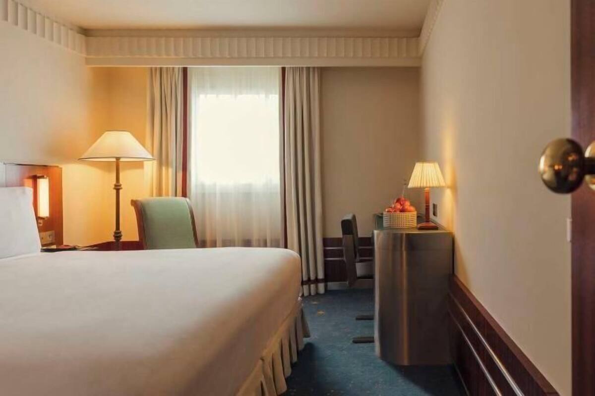 Quarto de hotel com cama, escrivaninha, cadeira, luminária e mesinha de centro com cesto.