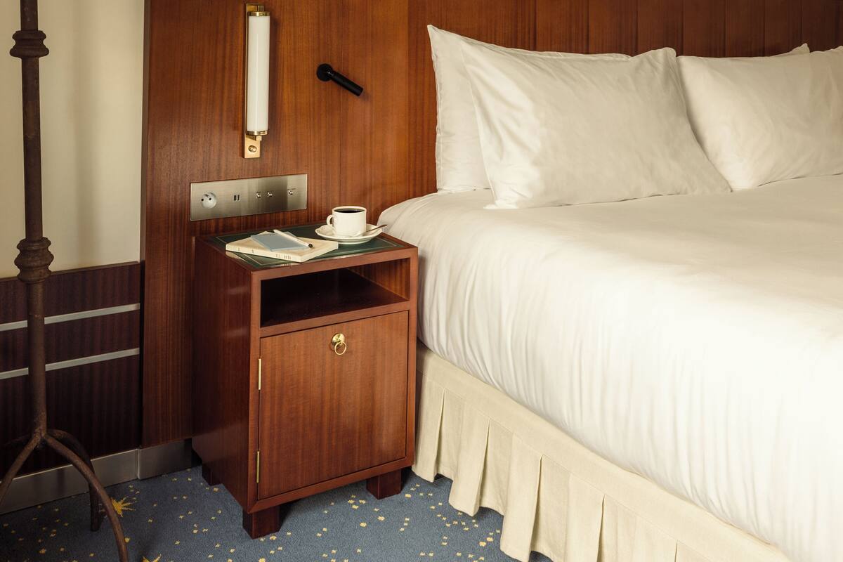 Quarto de hotel com cabeceira de madeira, mesa de cabeceira com luminária, quadro na parede e cama com roupa de cama branca.
