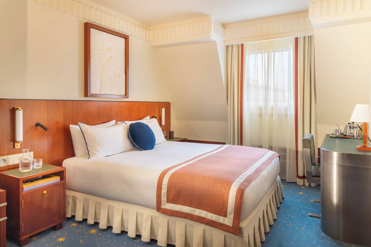 Quarto de hotel com uma cama grande, mesa de cabeceira com abajur, pequena escrivaninha e janela com cortinas.