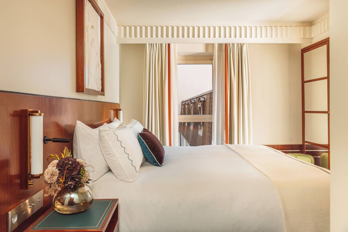 Quarto de hotel com uma cama grande, uma mesa de cabeceira com um vaso de flores, cabeceira de madeira e vista para uma varanda com cortinas.