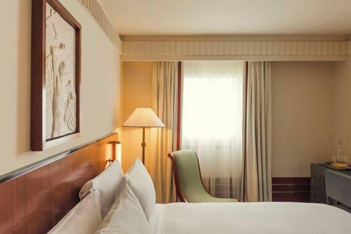 Quarto de hotel com cama, uma cadeira, luminária, obra de arte emoldurada e janela com cortinas.
