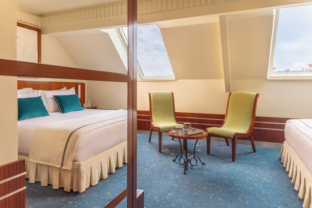 Quarto de hotel com beliche, duas cadeiras, uma mesinha e um claraboia.