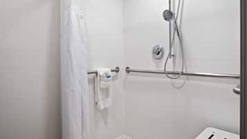 Um espaço de chuveiro pequeno, revestido com azulejos brancos, com um chuveiro manual, um porta-toalhas com toalha e um banco branco.