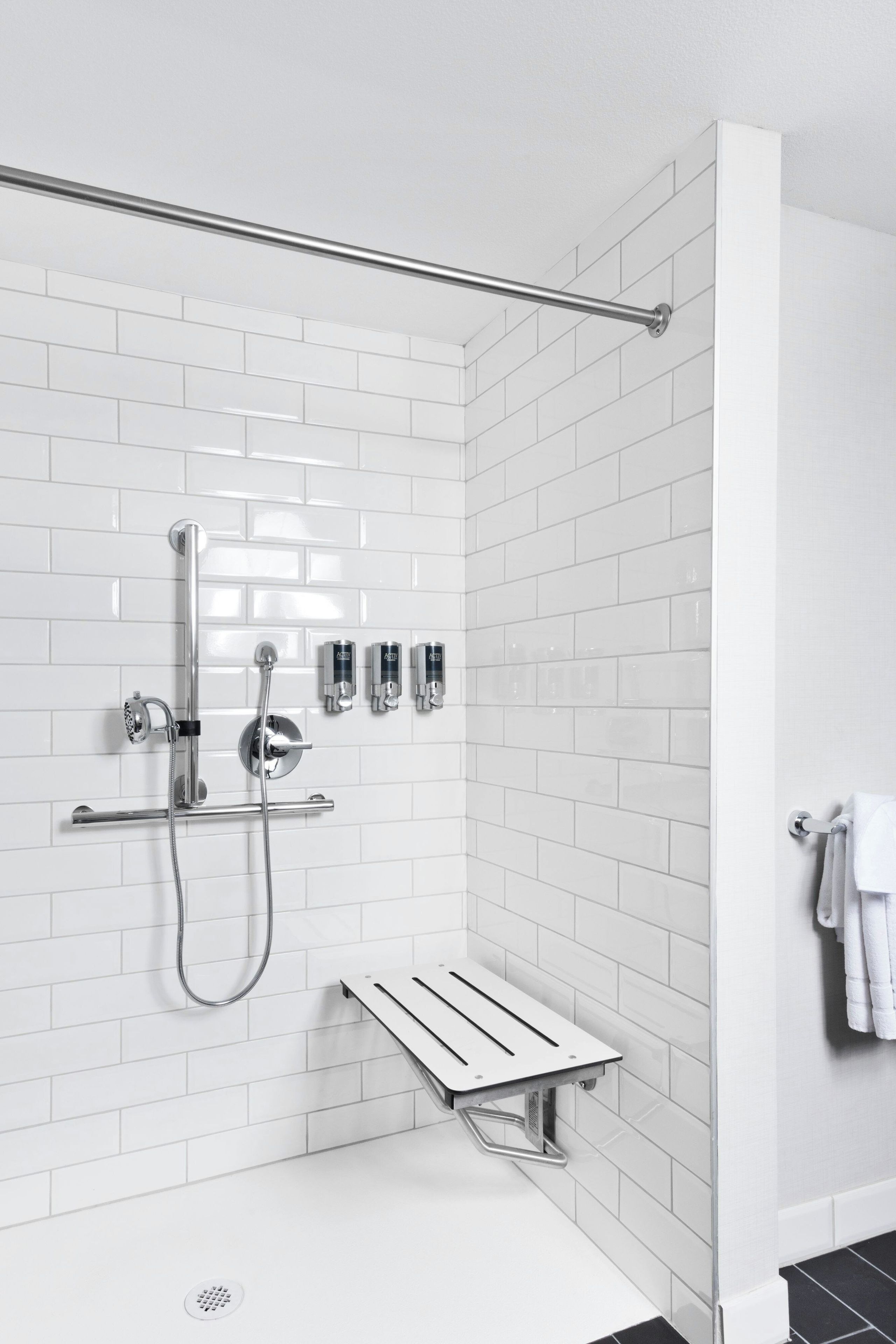 Casa de banho moderna com paredes revestidas a azulejos brancos, chuveiro manual, pulverizador de mão e um porta-toalhas com uma toalha dobrada.