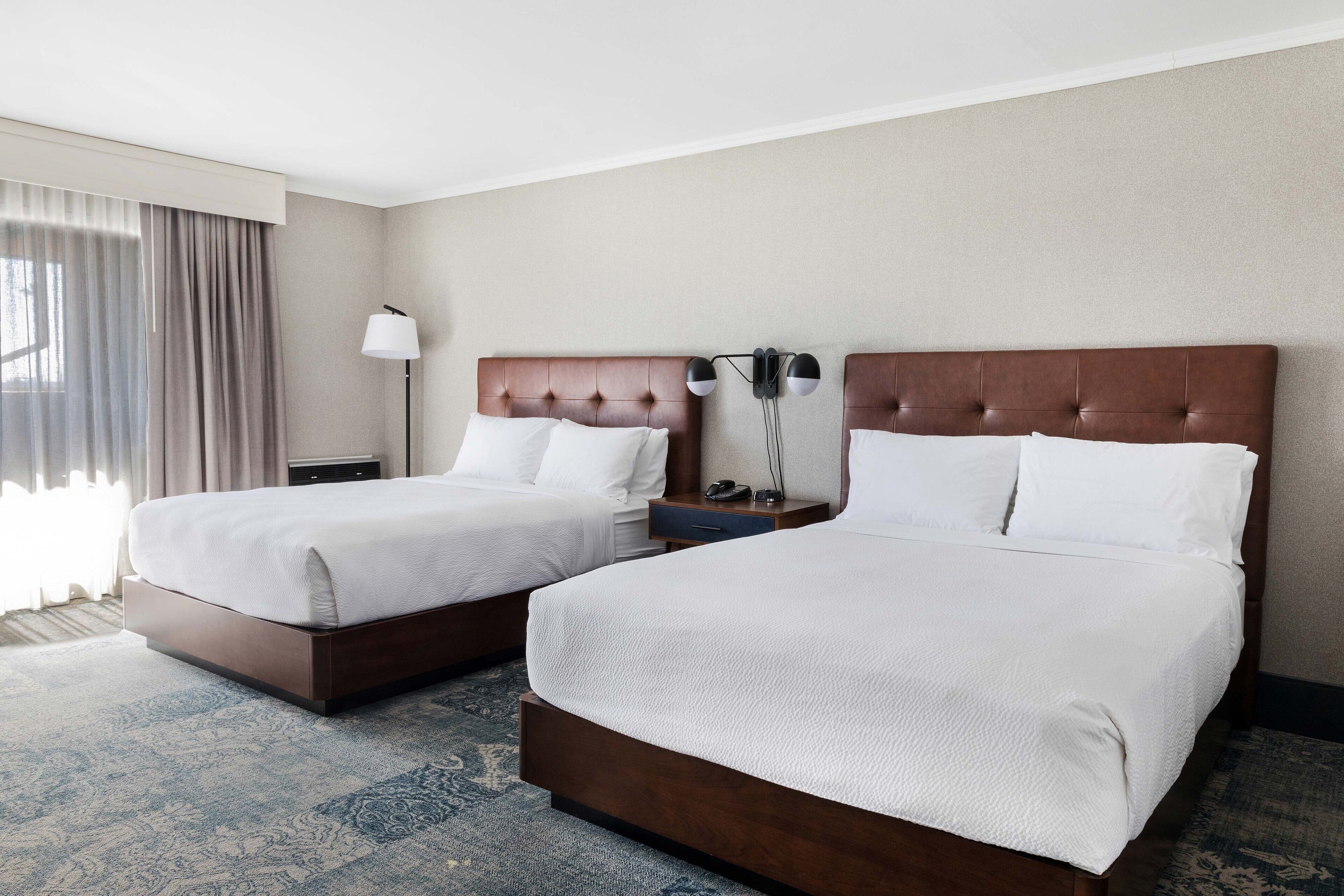 Quarto de hotel com duas camas, mesa de cabeceira, abajur e janela com cortinas.