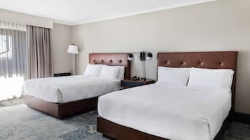 Quarto de hotel com duas camas, mesa de cabeceira, abajur e janela com cortinas.