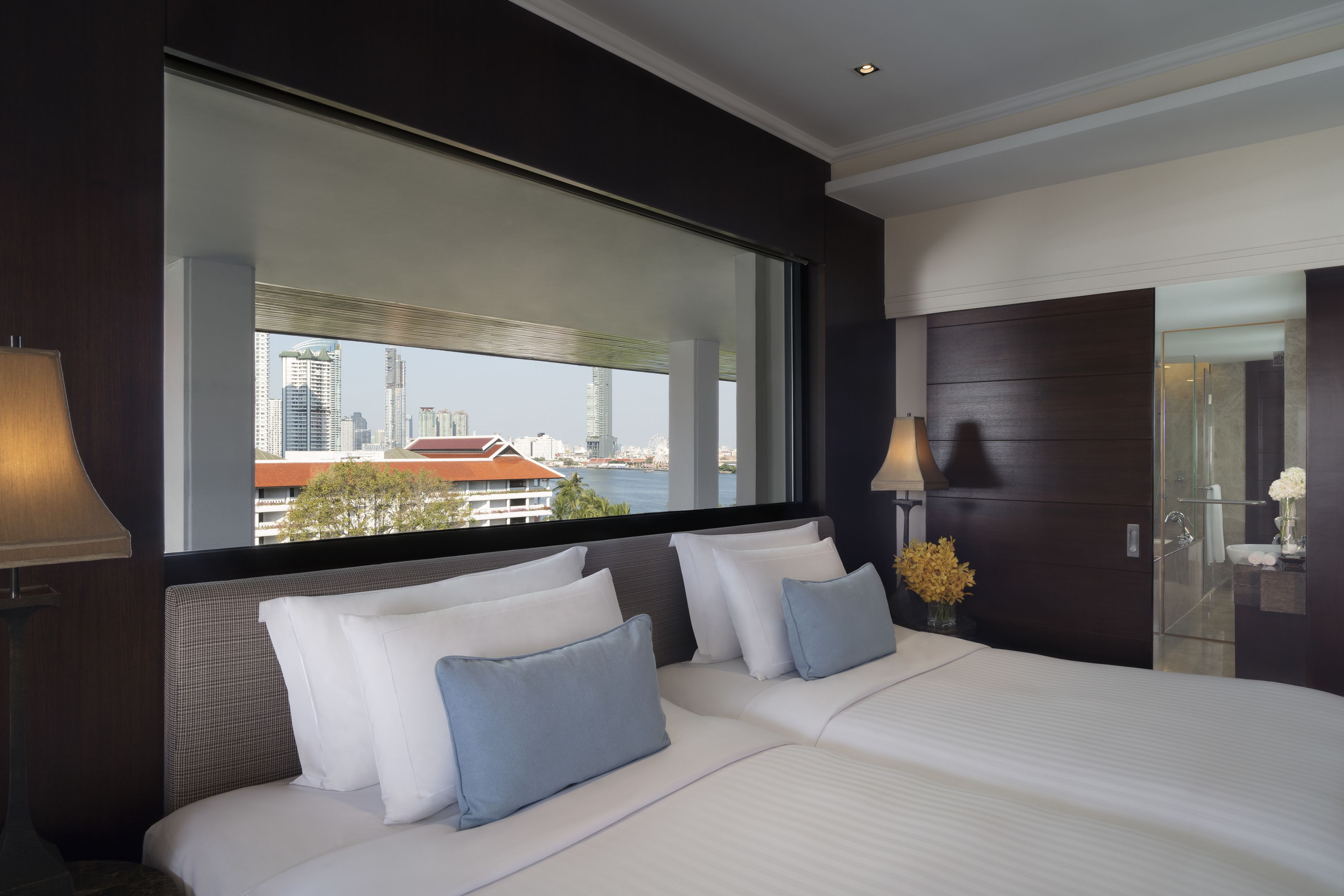 Quarto de hotel moderno com uma cama grande, duas luminárias de cabeceira, um criado-mudo e vista para a paisagem urbana através de uma janela ampla.