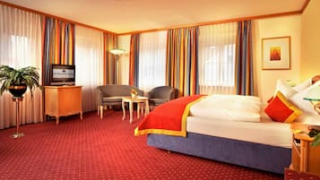 Quarto de hotel com uma cama, duas poltronas, uma mesinha, televisão e um vaso de planta.