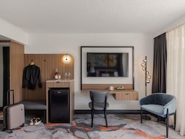 Quarto de hotel com escrivaninha, cadeira, minibar e televisão fixada na parede.
