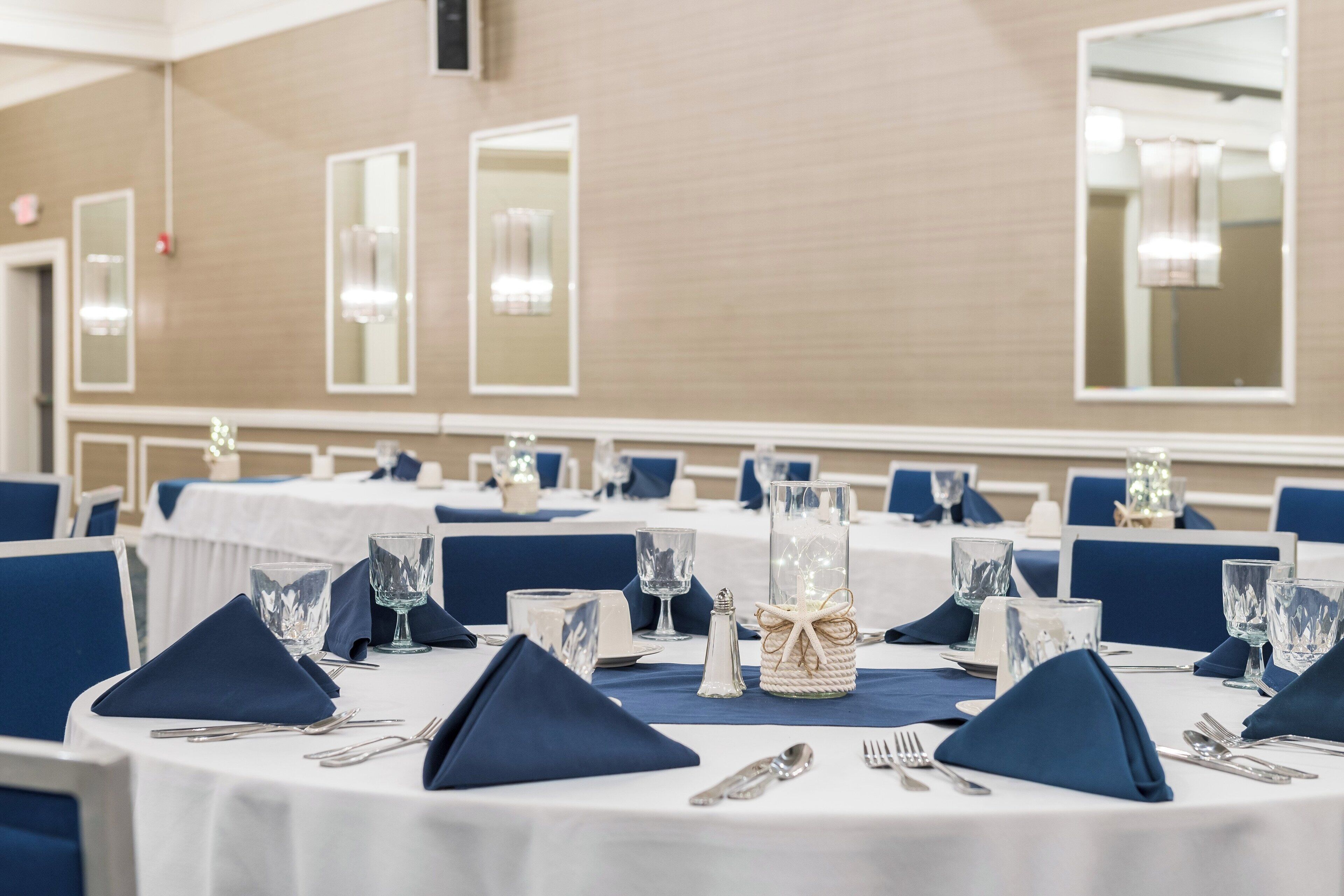 Sala de conferências com mesas redondas preparadas para uma reunião. Cada mesa tem toalha branca, guardanapos azuis e copos de vidro transparente.