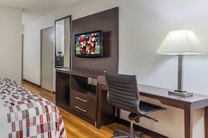 Quarto de hotel com cama, escrivaninha com cadeira, televisão fixada na parede e abajur na escrivaninha.