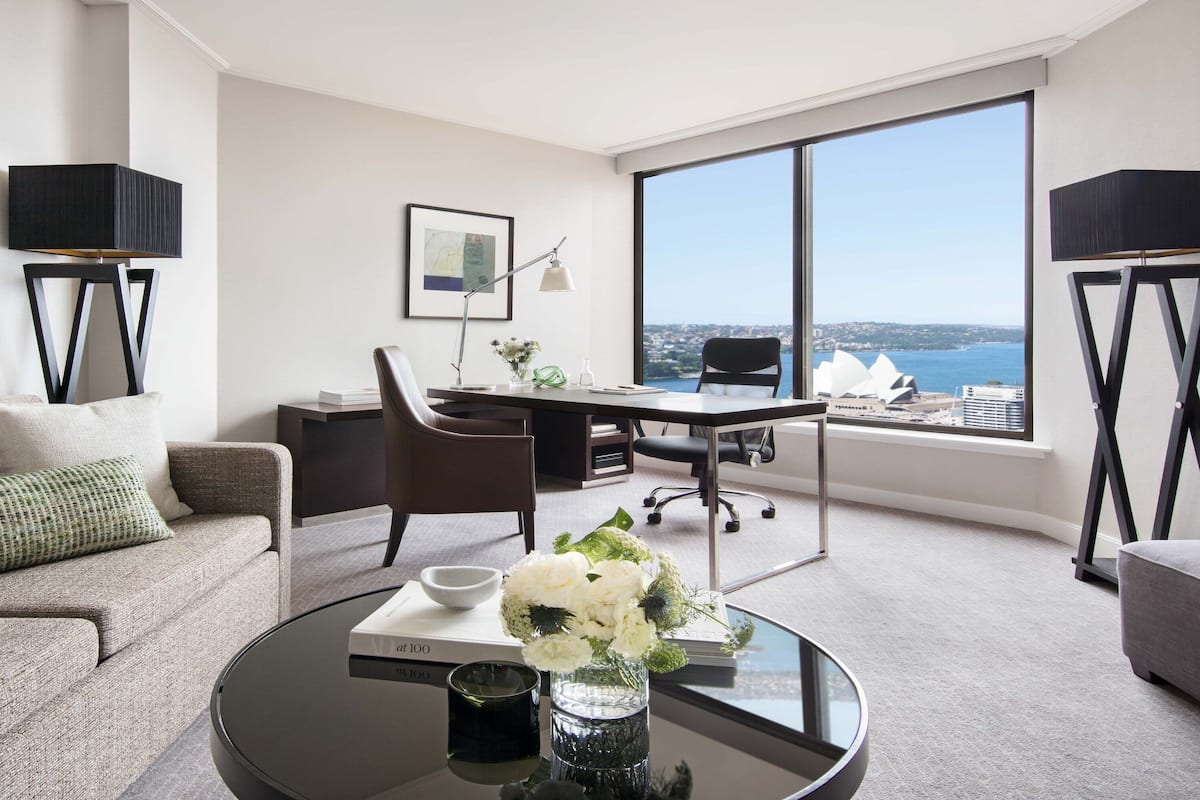 Quarto de hotel moderno com sofá, escrivaninha, cadeira, mesa de centro com um livro e flores, e uma janela grande com vista para a cidade.